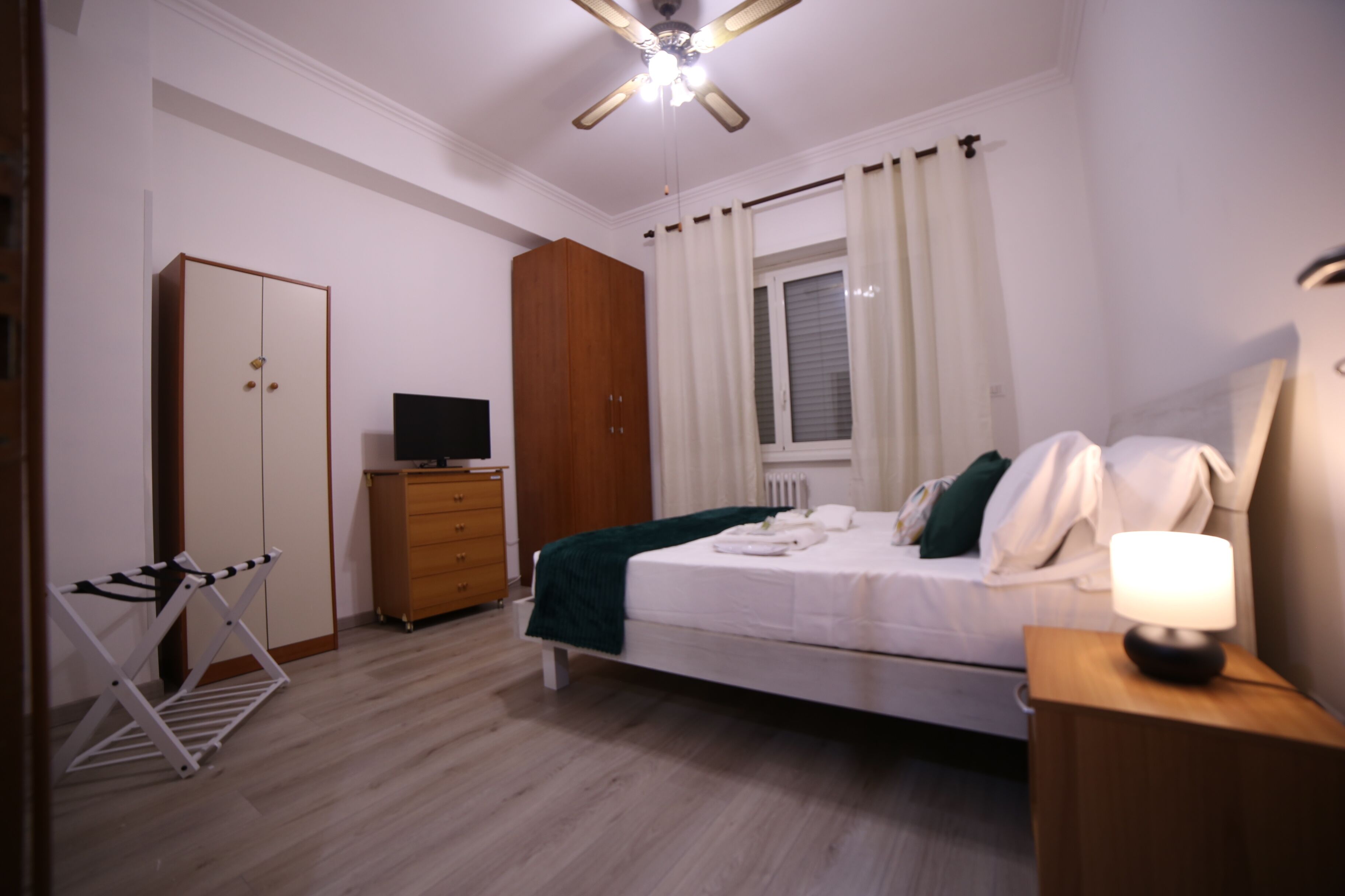 Quarto com cama, guarda-roupa de madeira, televisão sobre uma cômoda e ventilador de teto.