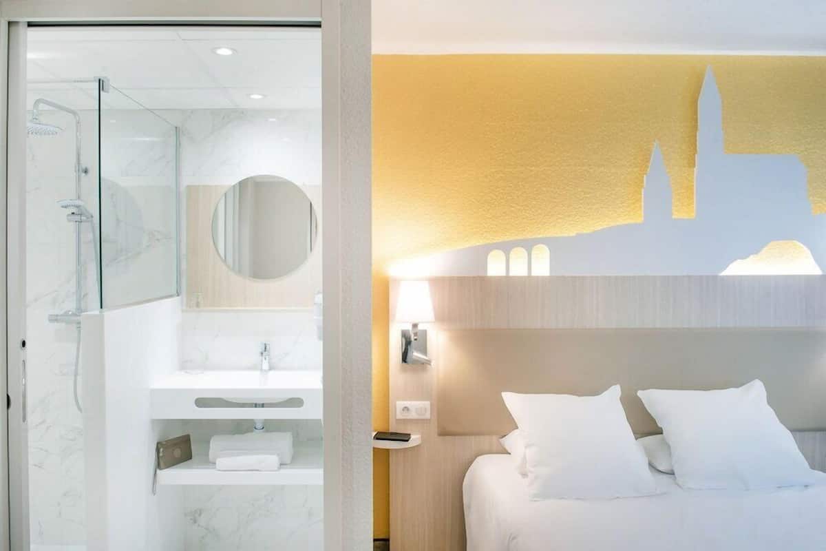 Quarto de hotel com box de vidro, pia branca, espelho, cama com roupa de cama branca e luminária fixada na parede.