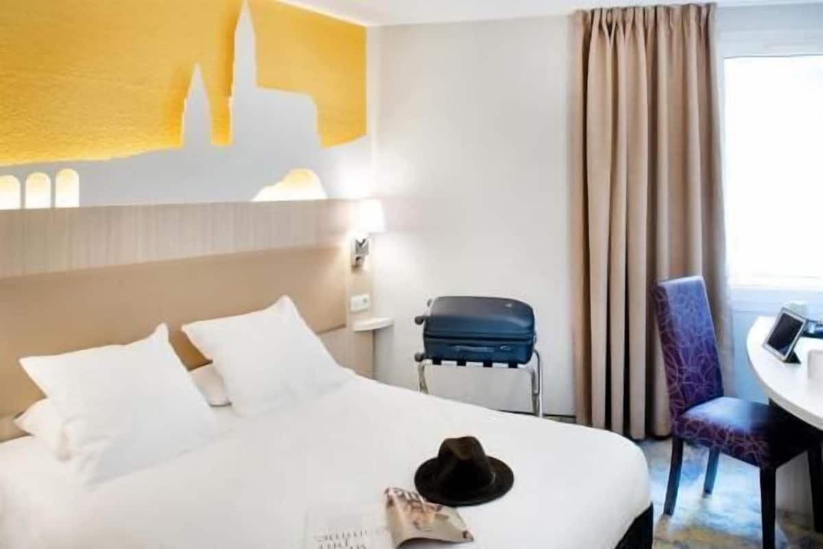 Quarto de hotel com cama, mala, chapéu, cadeira e mesa.