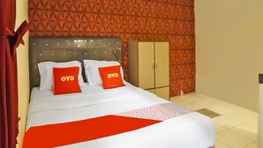 Quarto de hotel com uma cama, duas almofadas vermelhas com a marca 'OYO', um guarda-roupa e parede com padrão geométrico.