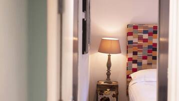 Um corredor estreito com uma cama à direita, um abajur sobre uma mesinha e um edredom colorido na cama.