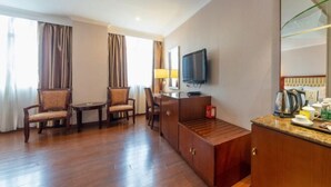 Quarto de hotel com piso de madeira, duas poltronas, uma mesa pequena, televisão, armário com espelho e um cantinho para café.