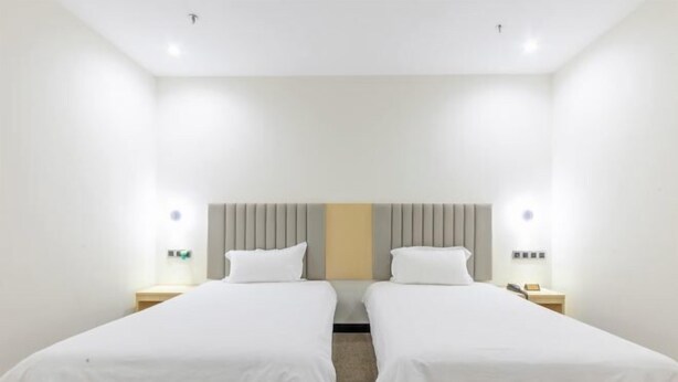 Quarto de hotel com duas camas de solteiro, cabeceira em tons neutros, mesas de cabeceira com abajures e uma parede branca lisa.
