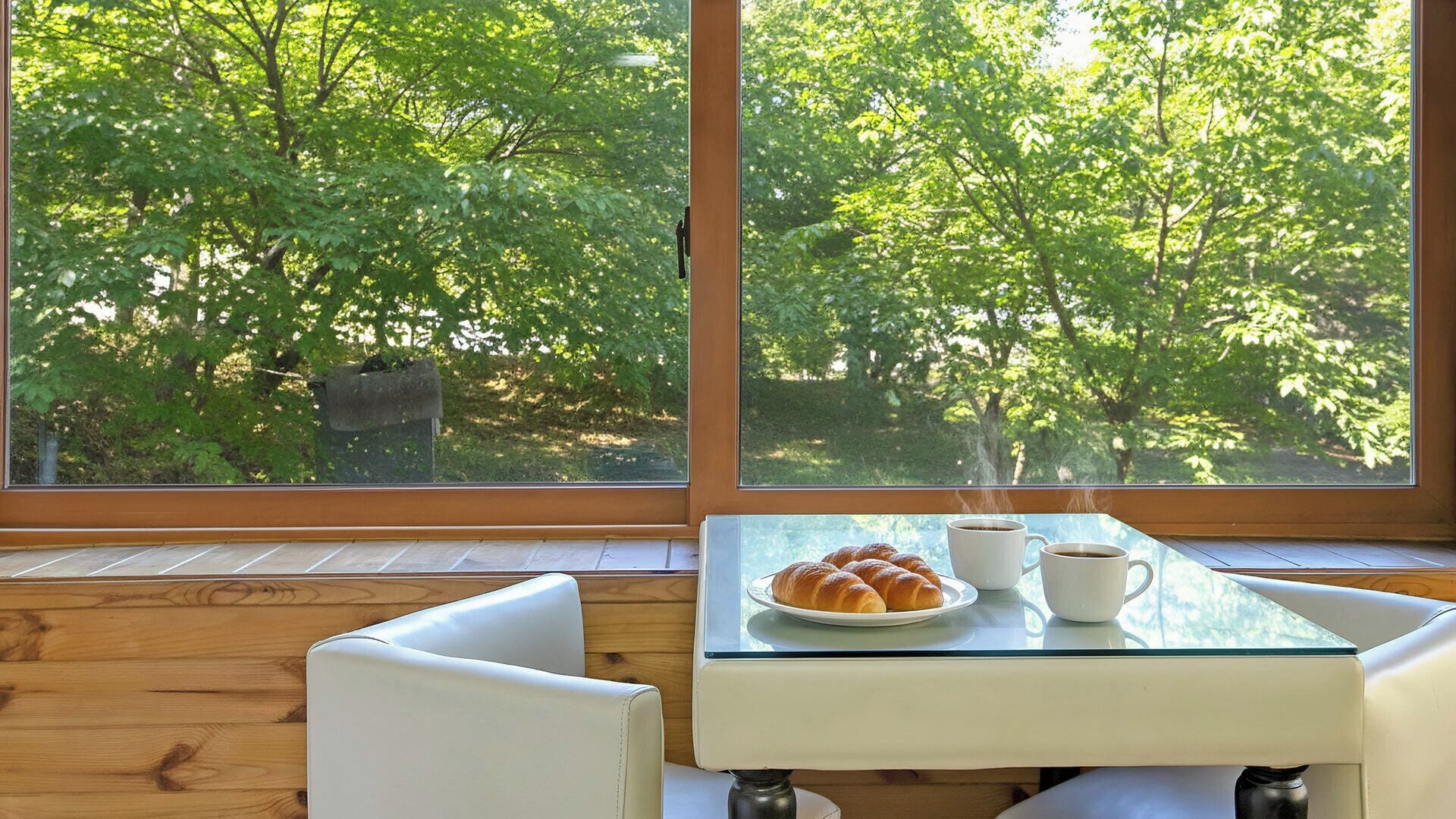 Quarto com vista para árvores através de grandes janelas, uma mesa com croissants e xícaras de café, e uma cadeira branca.