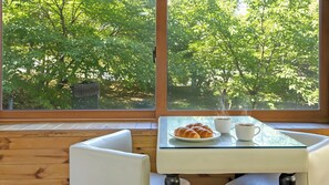 Quarto com vista para árvores através de grandes janelas, uma mesa com croissants e xícaras de café, e uma cadeira branca.