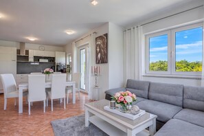 Uma sala de estar moderna com um sofá cinzento, uma mesa de jantar com cadeiras e uma cozinha ao fundo.