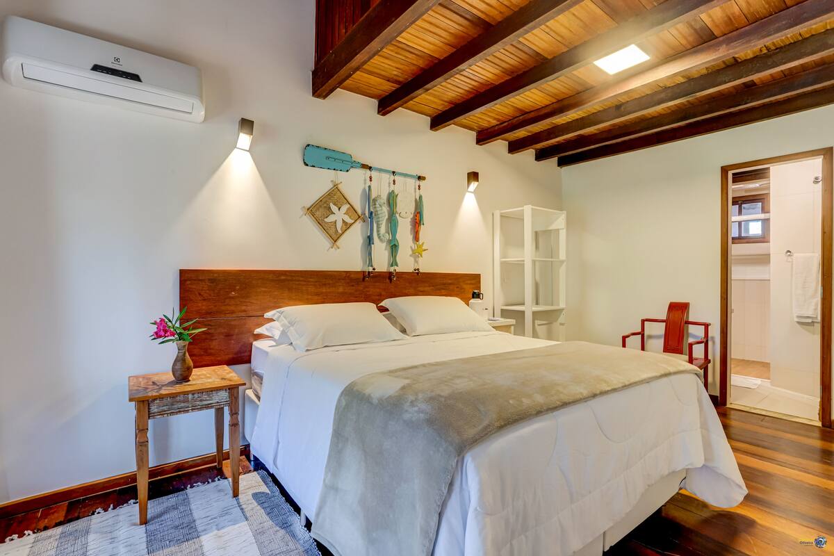 Quarto com cabeceira de madeira, mesa de cabeceira com um vaso, prateleira na parede e porta que dá para outro cômodo.