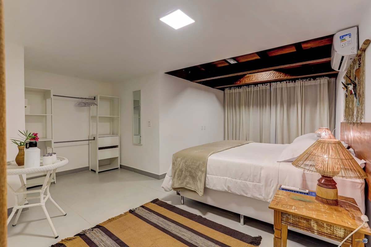 Quarto com cama, mesa de cabeceira, abajur e uma pequena mesa com um vaso.