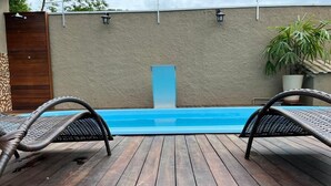 Um deck de madeira com piscina, duas cadeiras de vime, uma planta em vaso e uma parede com porta e duas luzes fixadas.