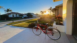 Uma bicicleta vermelha estacionada em um caminho de paralelepípedos, com vista para uma rua residencial e casas ao fundo.