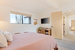 Quarto com cama grande, televisão, janela vista para árvores e porta para outro cômodo.