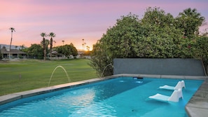 Piscina com vista para o pôr do sol, área gramada e uma casa ao fundo.