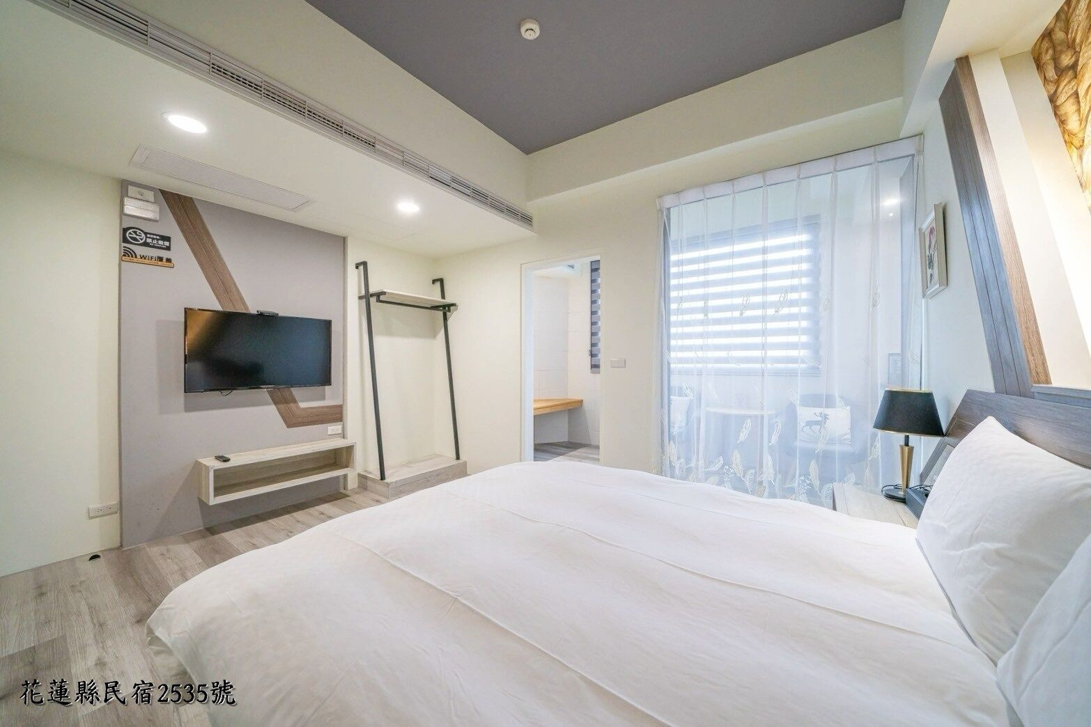 Quarto de hotel moderno com uma cama grande, uma televisão de tela plana na parede, uma pequena escrivaninha com abajur e uma janela com persianas.