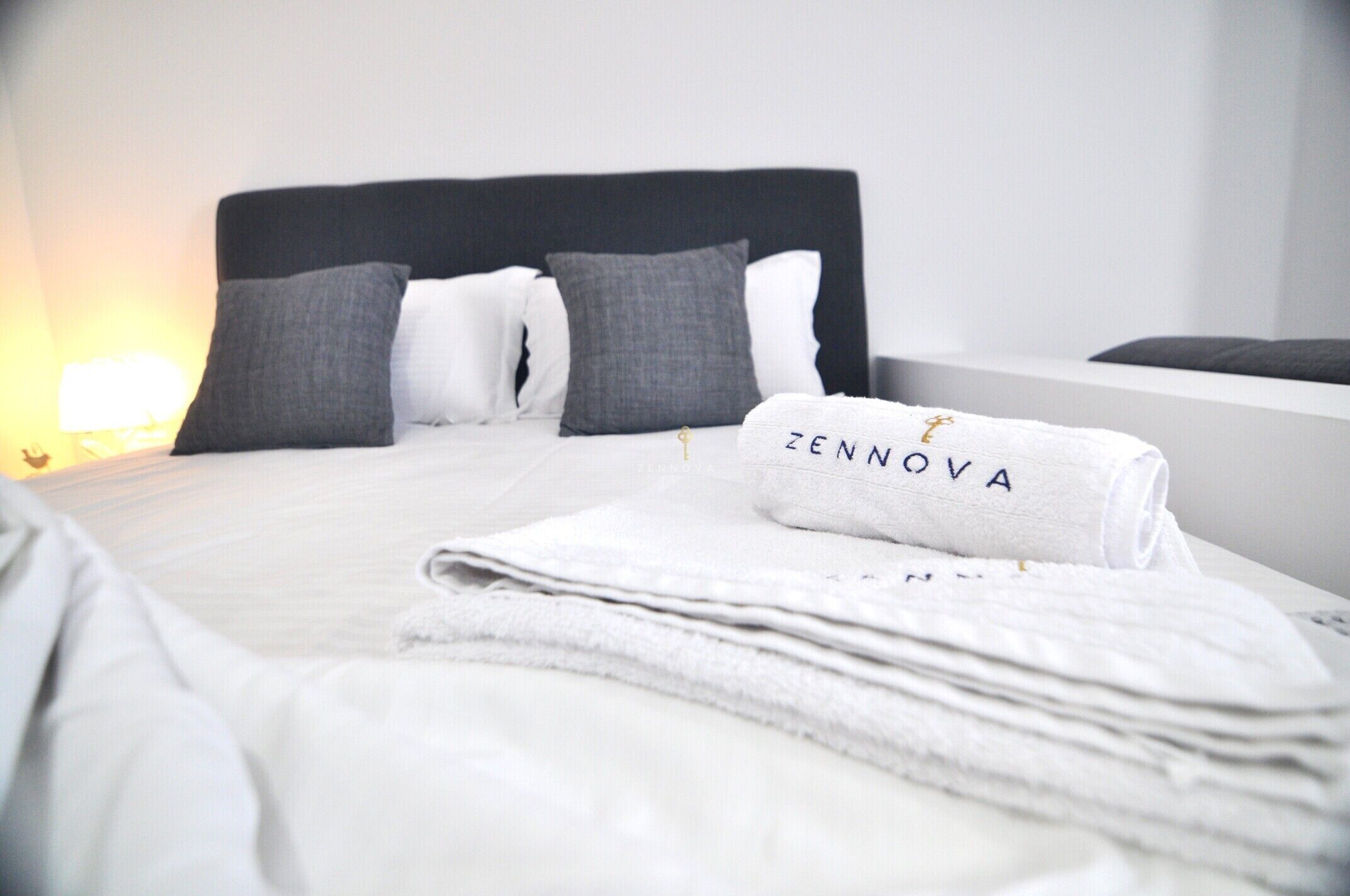 Cama bem arrumada com uma toalha dobrada com a etiqueta "ZENOVA" e uma pilha de toalhas brancas.