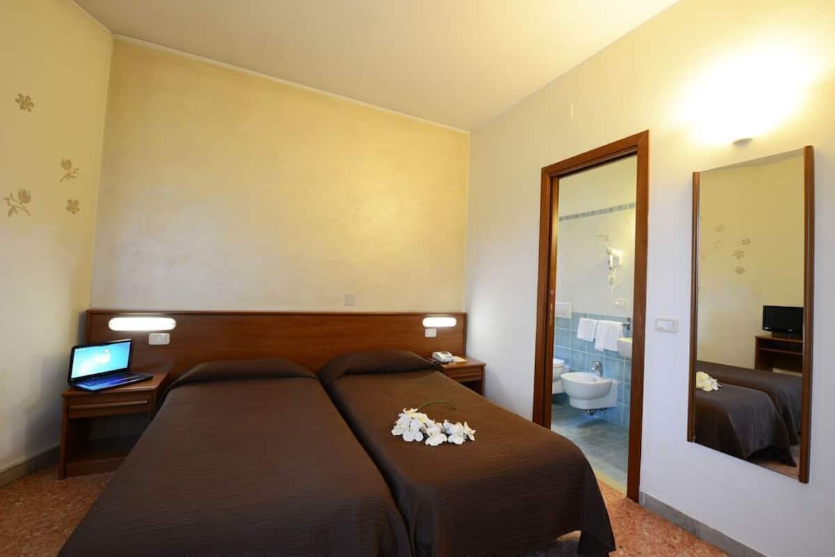 Quarto de hotel com duas camas, uma mesa com um laptop, mesa de cabeceira com telefone, banheiro com pia e vaso sanitário, e um espelho na parede.