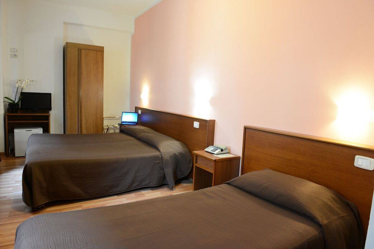 Quarto de hotel com duas camas, um guarda-roupa de madeira, uma escrivaninha com computador e uma mesinha com telefone.