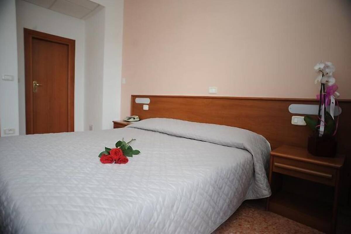 Uma cama bem arrumada com colcha branca, cabeceira de madeira e mesa de cabeceira com um vaso de flores.
