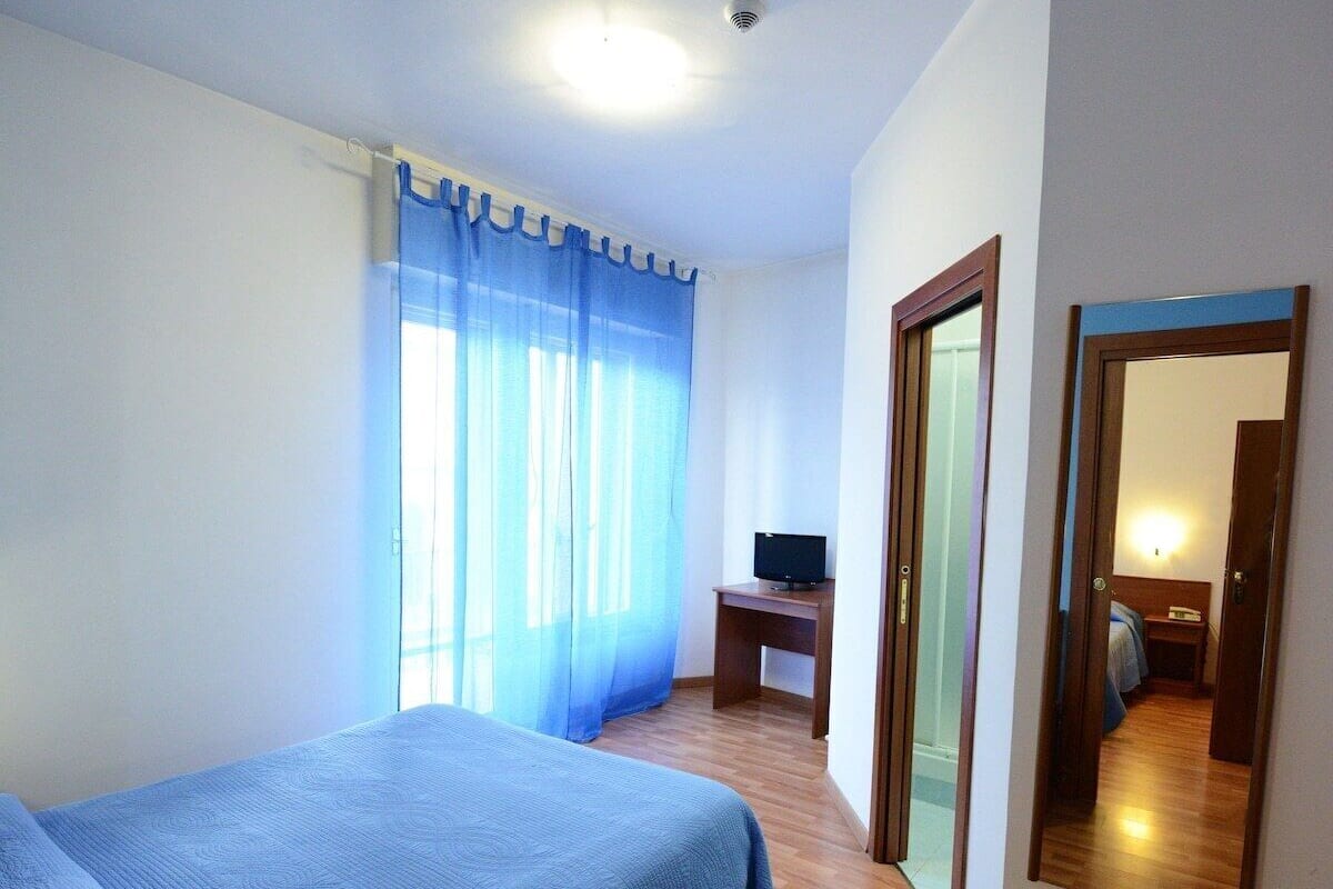 Quarto de hotel com duas camas, uma escrivaninha, uma cadeira, um espelho e uma luminária no teto.