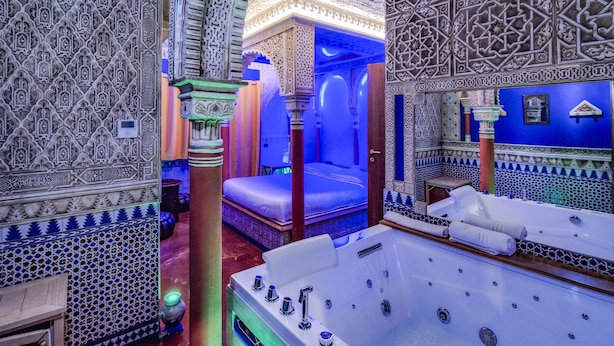 Uma casa de banho luxuosa com trabalhos em azulejos detalhados, uma banheira grande e uma cama com dossel iluminado em azul.