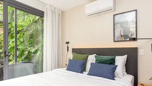 Quarto de hotel com cama, mesa de cabeceira, ar condicionado, janela com cortinas e vista para o verde lá fora.