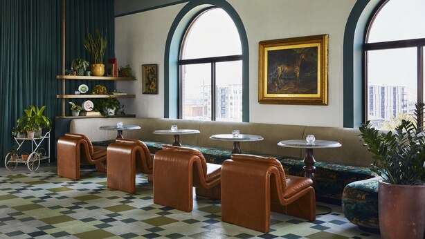 Quarto com piso quadriculado, poltronas de couro, um sofá e uma pequena mesa com um copo. Há janelas com arcos e vista para edifícios.
