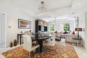 Uma sala de estar ampla com mesa de jantar, assentos confortáveis, um candelabro e uma grande janela que oferece iluminação natural.
