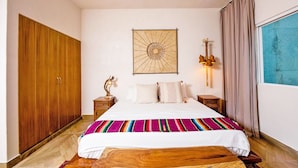 Quarto com uma cama grande, um guarda-roupa de madeira, uma obra de arte na parede, uma mesinha de cabeceira com abajur e uma janela com cortinas.