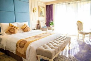 Quarto de hotel com uma cama grande, área de estar com uma poltrona e uma mesinha, e janela com cortinas.