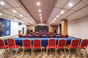 Sala de conferências com uma mesa comprida, cadeiras vermelhas e um palco na frente.