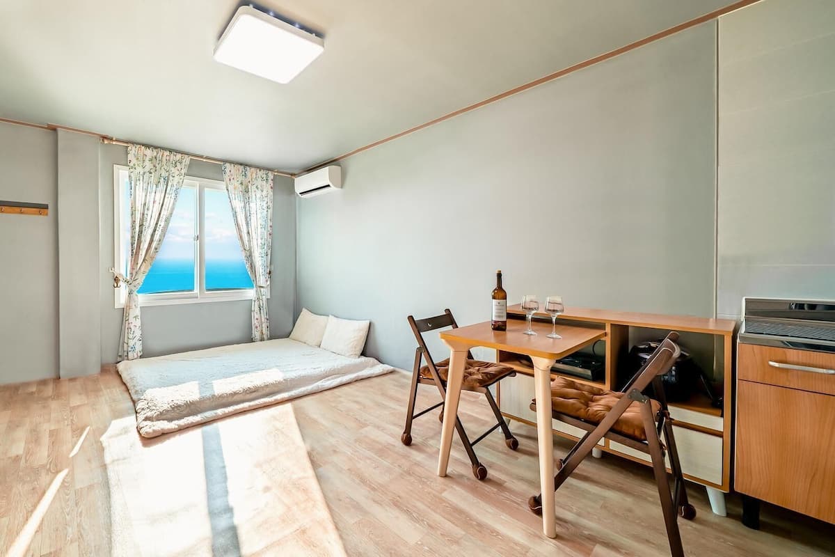Quarto compacto com cama, escrivaninha com cadeira, televisão e janela com cortinas.