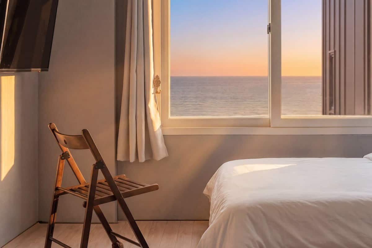 Quarto com cama, cadeira de madeira, janela com cortinas e vista para o mar.