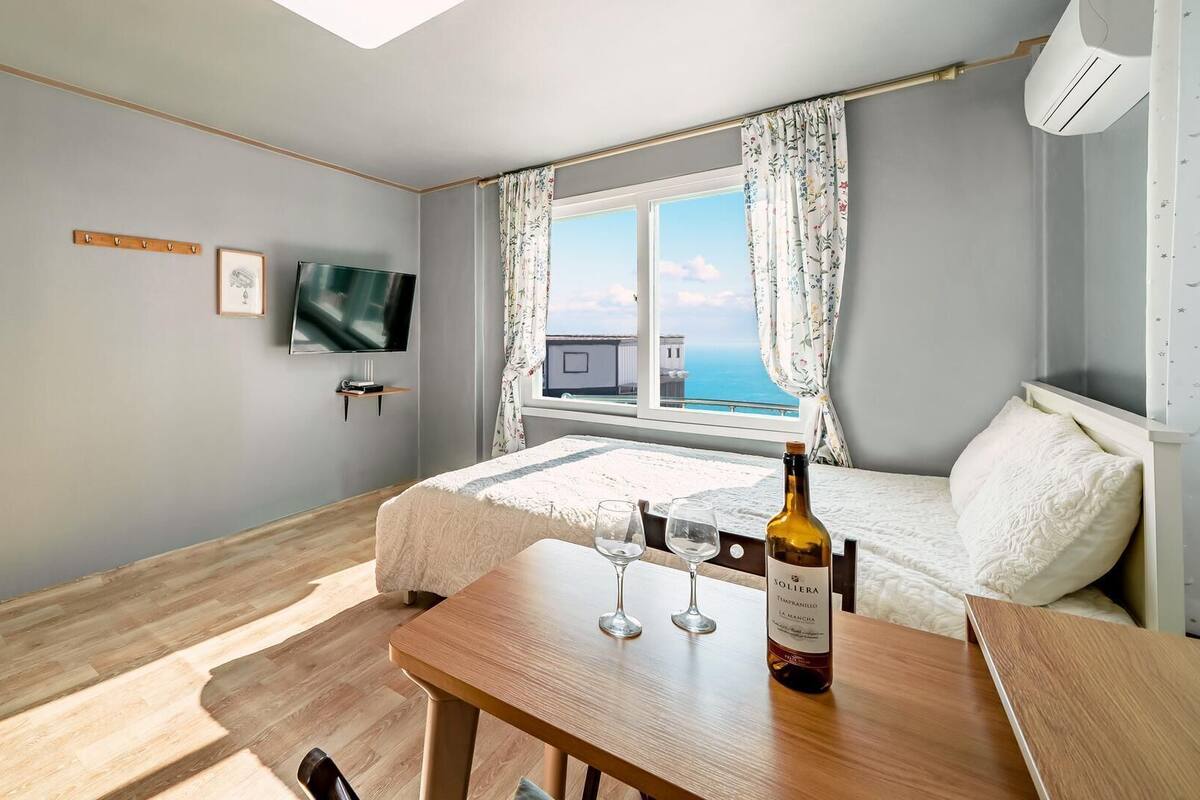 Quarto com cama, televisão, janela com cortinas, mesa com garrafa e copos, e piso de madeira.