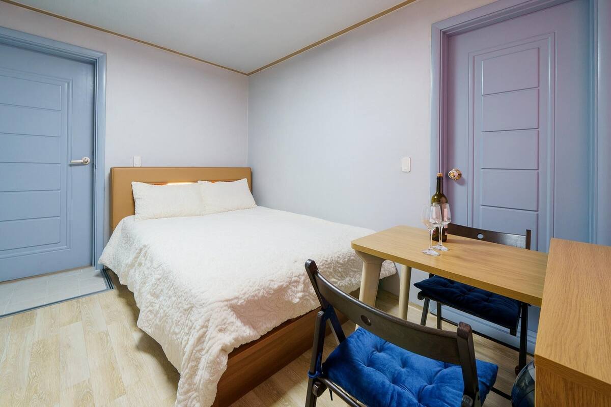 Quarto com cama, mesa de madeira e uma cadeira. Há uma garrafa de vinho sobre a mesa.