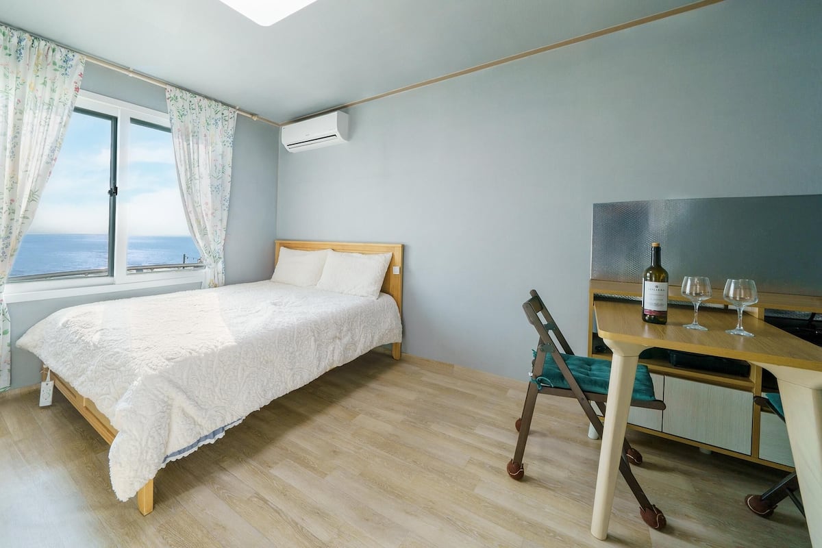 Quarto com cama, janela com cortinas, escrivaninha com cadeira, garrafa de vinho e copos.