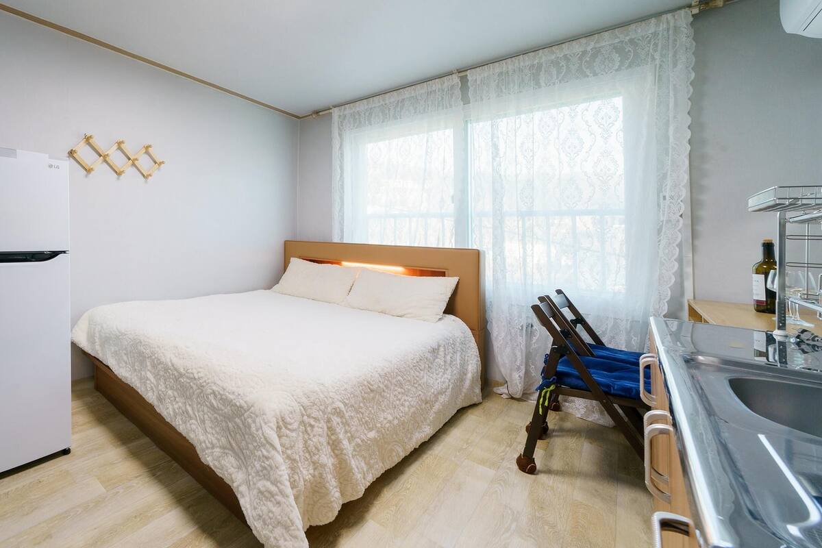 Quarto com cama, guarda-roupa branco, cadeira de madeira e uma área de cozinha com pia e garrafa de vinho.