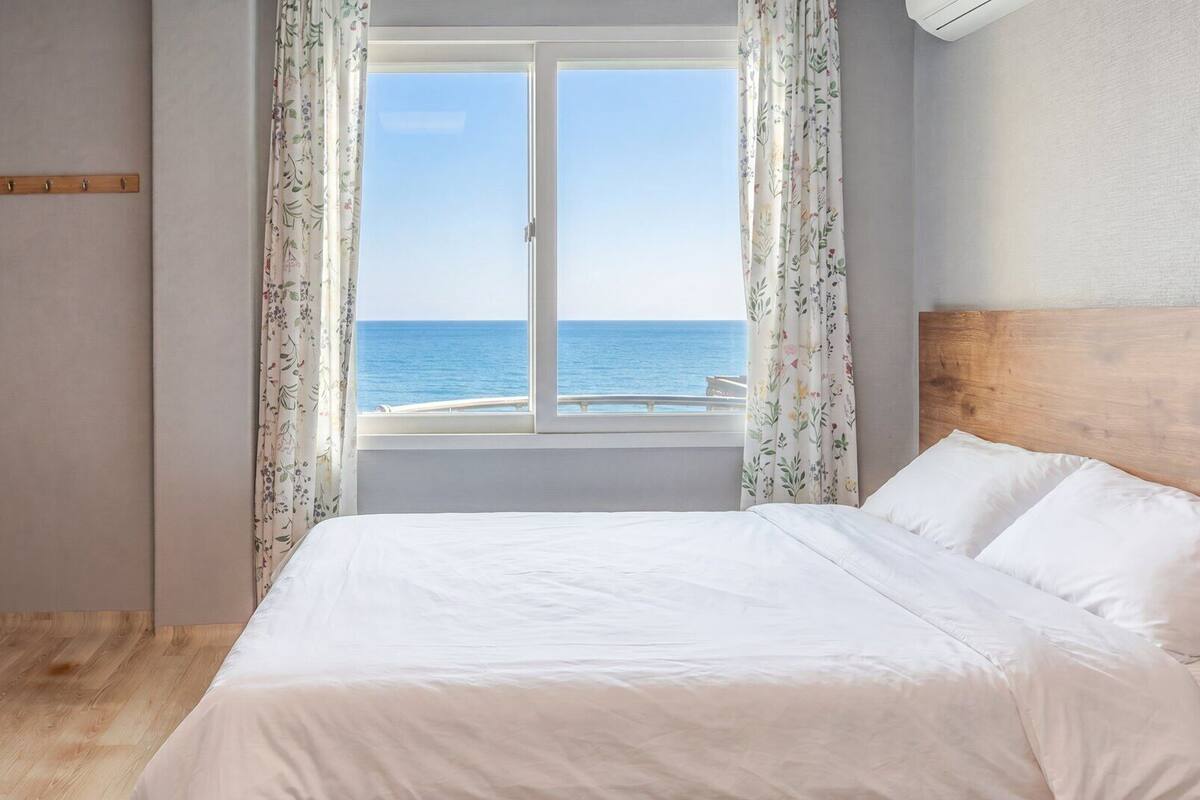 Quarto com cama, cabeceira de madeira, janela com cortinas e vista para o mar.