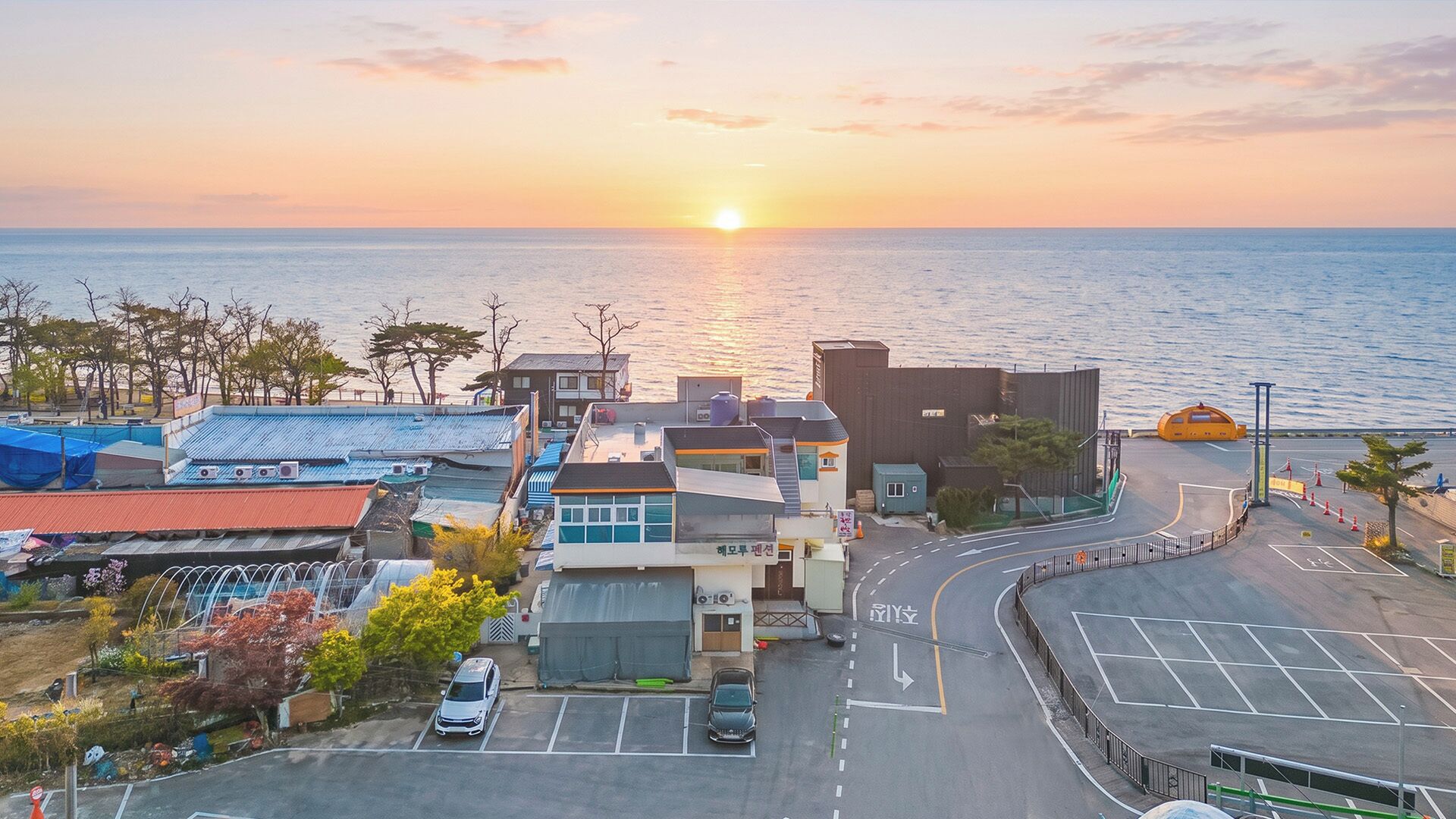 Uma vila costeira com área de estacionamento, edifícios e vista do pôr do sol sobre o mar.