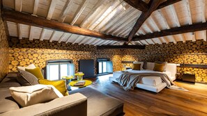 Um quarto espaçoso com teto alto, vigas de madeira aparentes, parede de pedra com padrão de troncos, uma cama grande, um sofá e uma mesa de centro.
