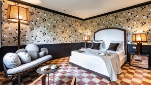 Quarto de hotel com chão xadrez, uma cama grande com cabeceira, duas mesas de cabeceira com abajures, um banco com almofadas e uma parede com papel de parede temático de cavalos.