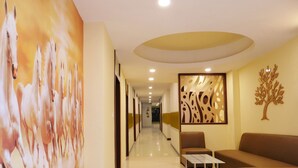 Um saguão de hotel com um corredor longo, uma área de recepção com um sofá e uma mesa pequena, e uma grande pintura abstrata na parede à esquerda.