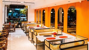 Interior de restaurante moderno com mesas grandes, cadeiras de vime e uma parede de destaque laranja vibrante.