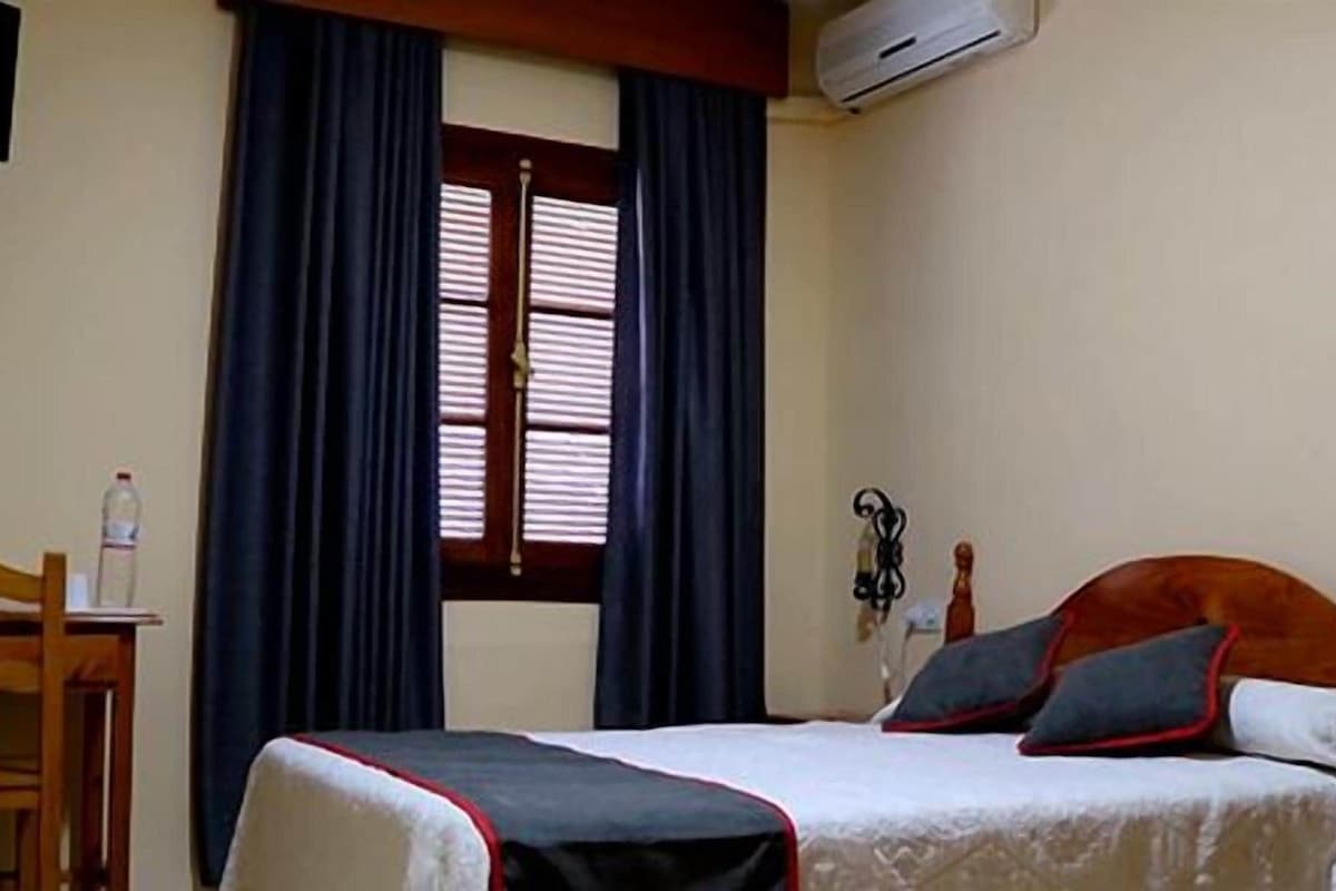 Quarto de hotel com cama, escrivaninha, cadeira, janela com cortinas e ar condicionado.