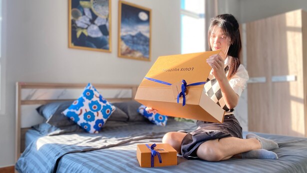 Uma pessoa sentada em uma cama com uma caixa Louis Vuitton e uma caixa de presente menor.