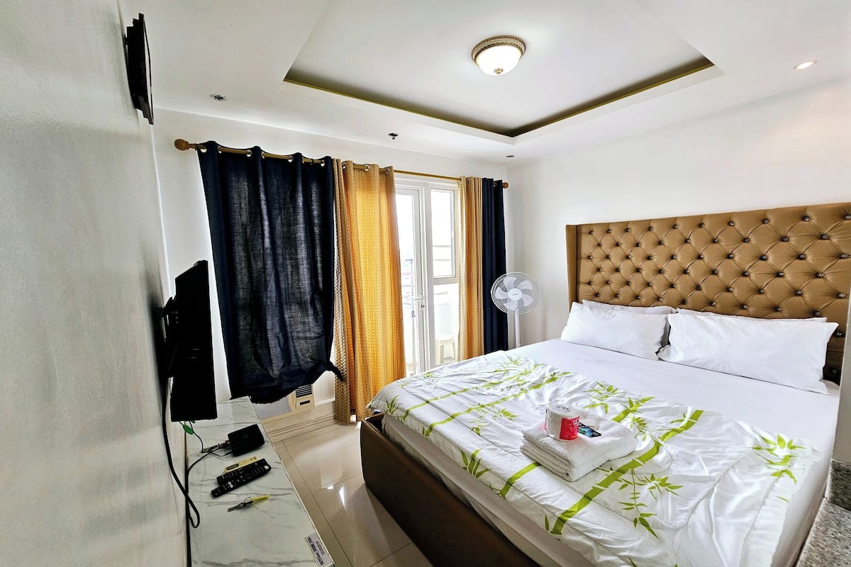 Quarto de hotel com uma cama grande, cabeceira estofada, janela com cortinas e ventilador de teto.