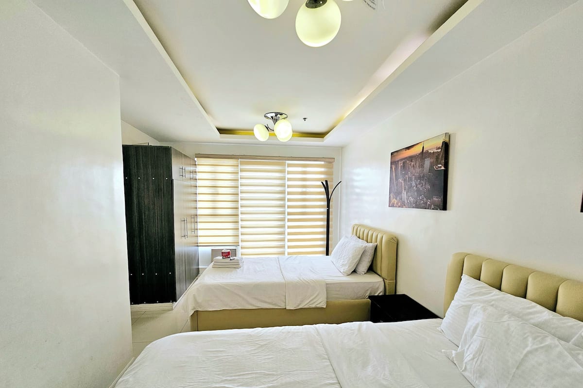 Quarto de hotel com duas camas, uma janela grande com persianas, um quadro na parede e um lustre.
