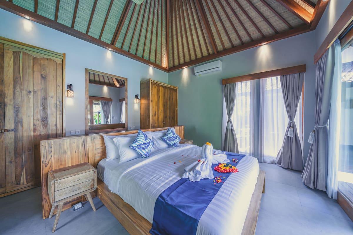 Quarto com cabeceira de madeira, mesa de cabeceira de madeira, cama com colcha azul e uma grande janela com cortinas.