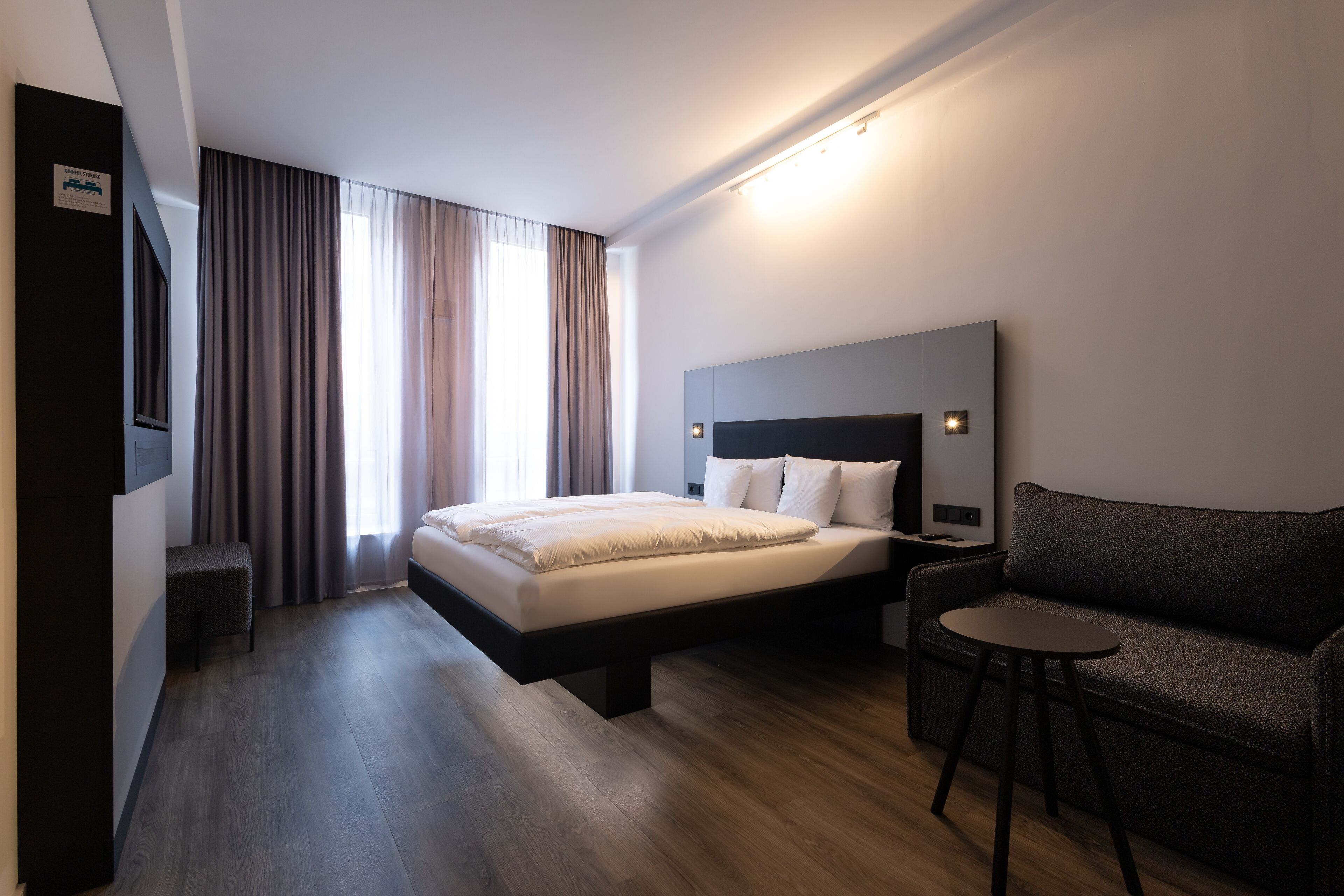 Quarto de hotel moderno com uma cama grande, um sofá, uma mesinha e uma TV fixada na parede.