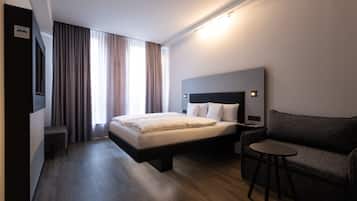 Quarto de hotel moderno com uma cama grande, um sofá, uma mesinha e uma TV fixada na parede.