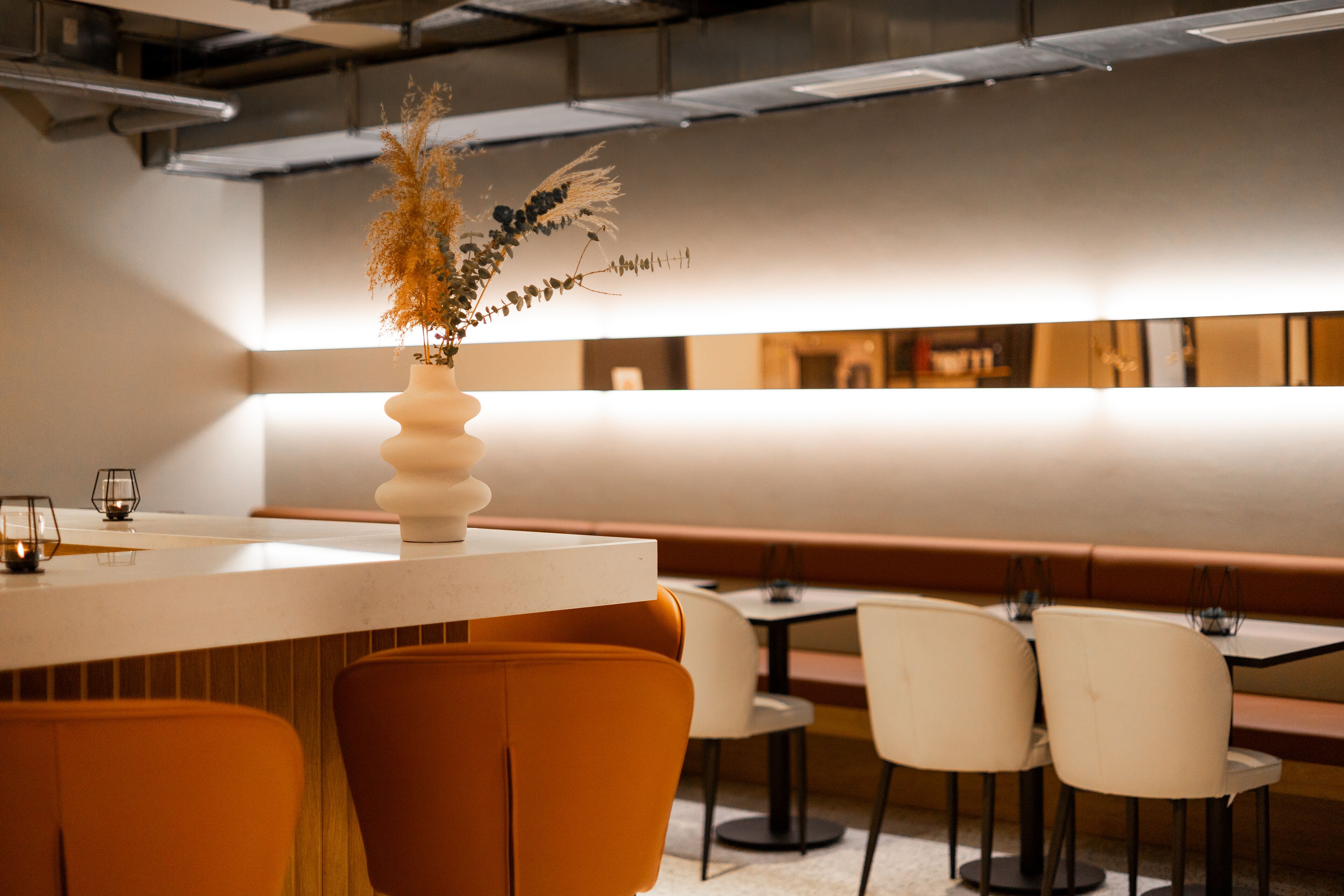 Interior de restaurante moderno com mesa branca, cadeiras laranja e um vaso com flores secas.