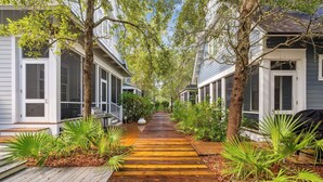 Uma passagem de madeira ladeada por casas com detalhes brancos e vegetação verde, que leva a uma área coberta.